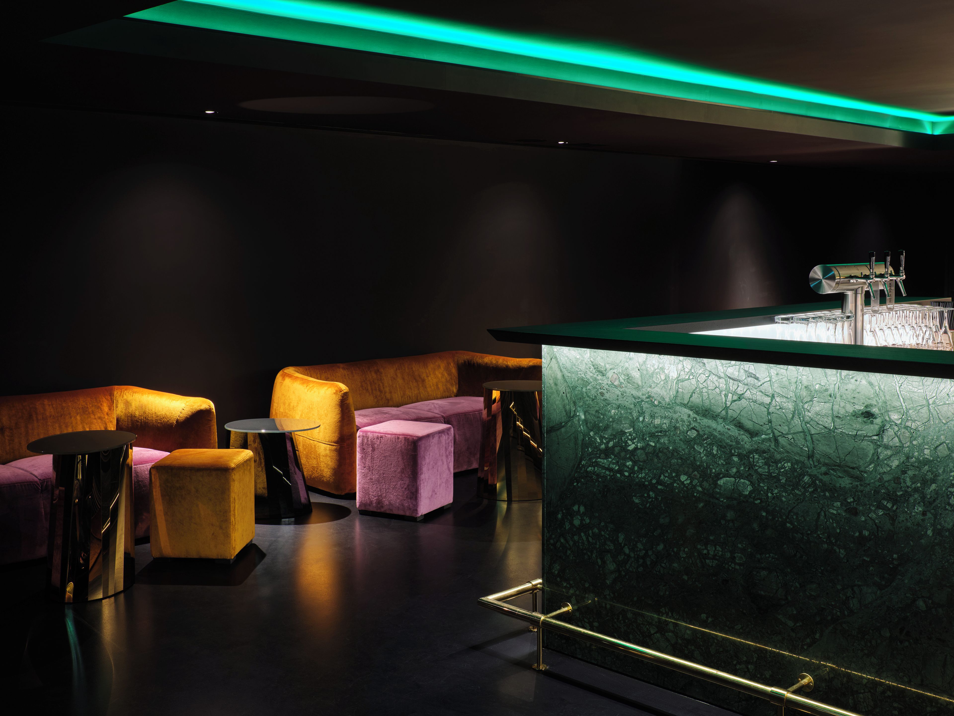 Uma área de bar com bancada de mármore, assentos acolchoados e teto iluminado em tons de verde.