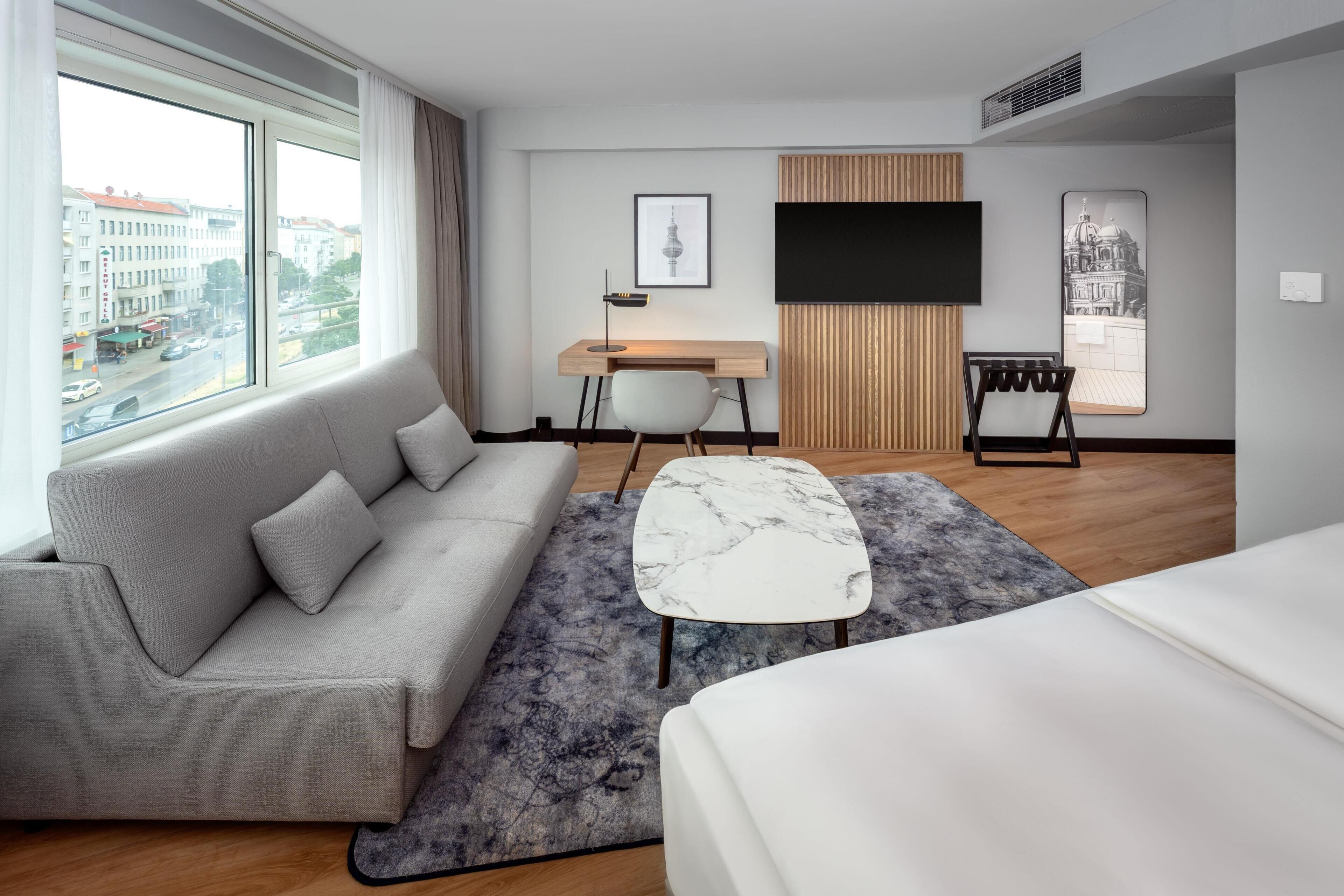 Quarto de hotel moderno com sofá, uma pequena mesa, escrivaninha, televisão e vista para a cidade.