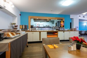 Espaço de café da manhã com serviço de buffet, área de estar com mesa e cozinha ao fundo.