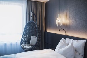 Quarto de hotel com uma cadeira suspensa, cama com roupa de cama branca, luminária fixada na parede e vista do exterior por uma janela com cortinas.