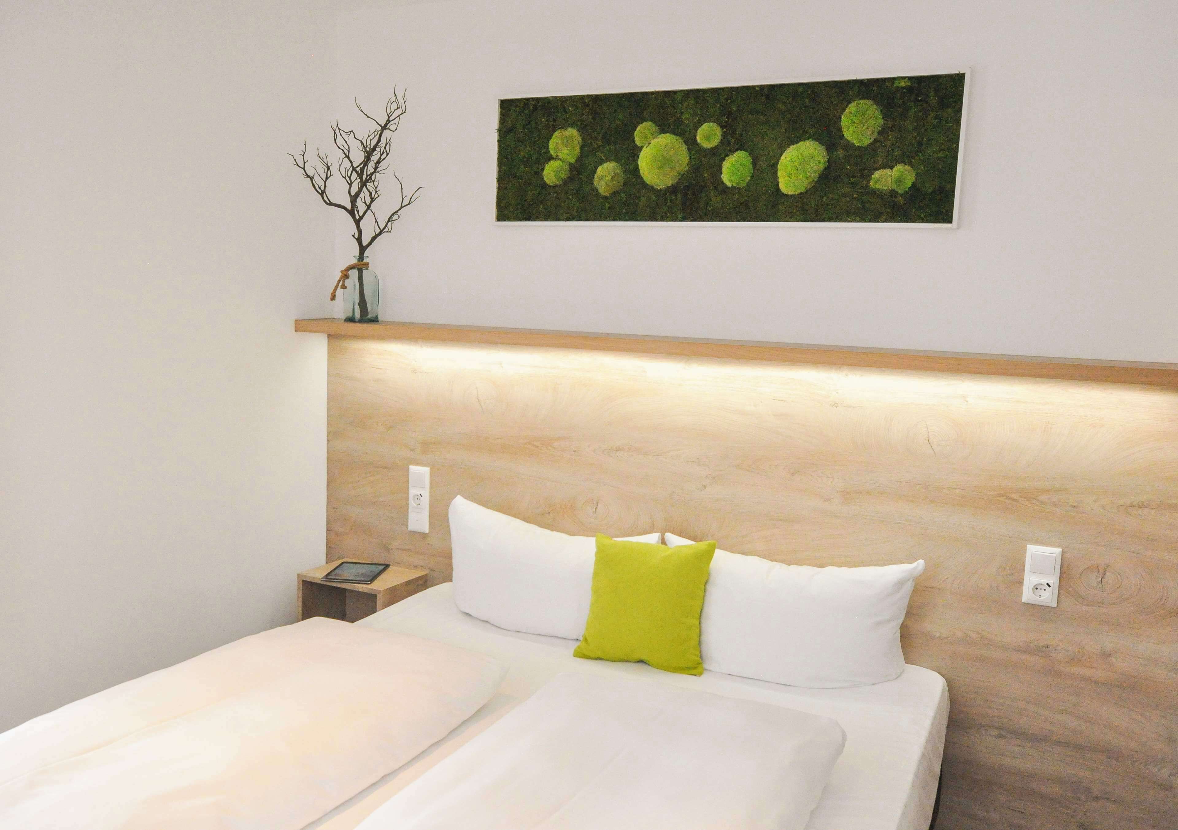 Quarto de hotel com cabeceira de madeira, duas camas, mesa de cabeceira com abajur, pequeno vaso com galho e pintura abstrata verde na parede.