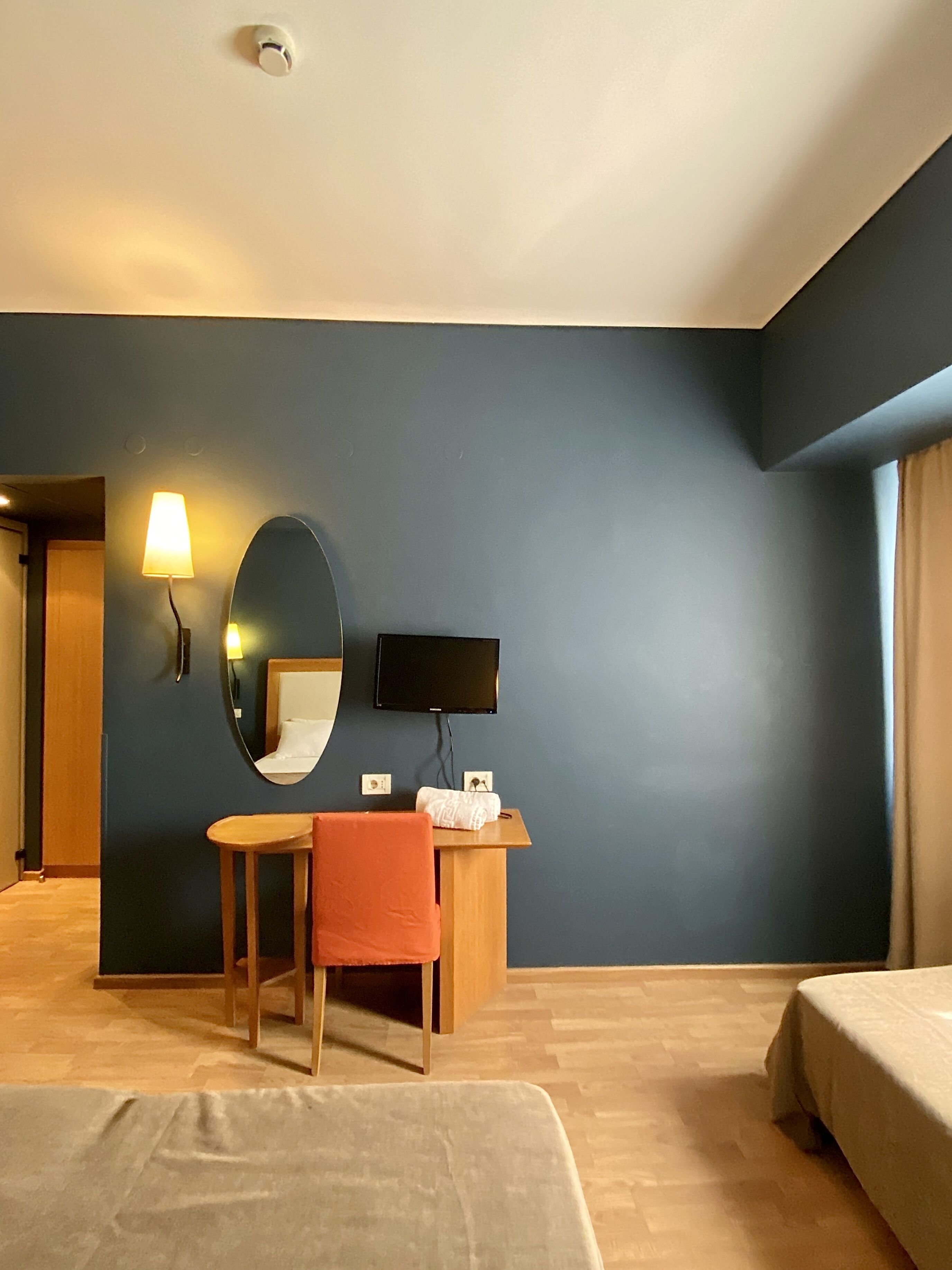 Quarto de hotel com cama, escrivaninha com cadeira, TV de tela plana, luminária de parede e espelho.