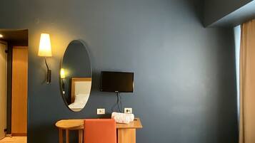 Quarto de hotel com cama, escrivaninha com cadeira, TV de tela plana, luminária de parede e espelho.