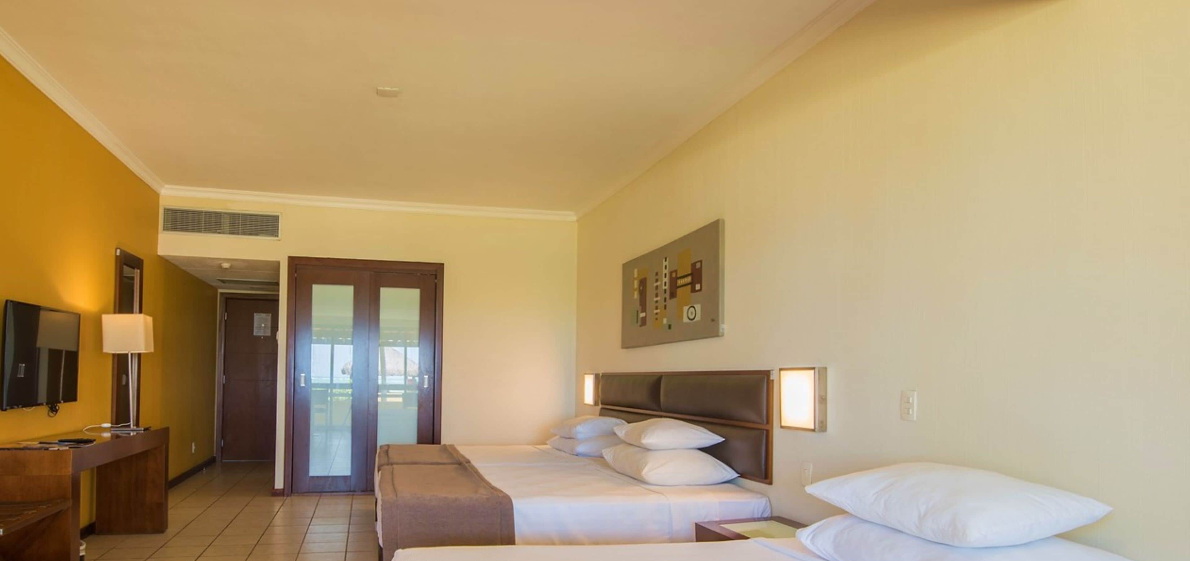 Quarto de hotel com duas camas, uma escrivaninha com luminária, televisão e porta para uma varanda.