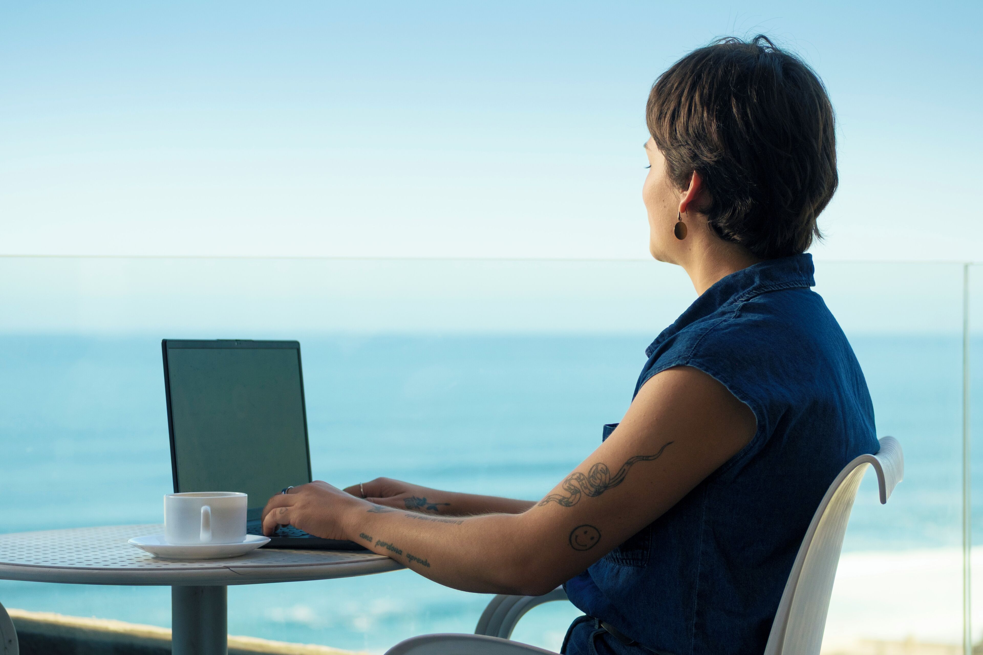 Uma pessoa à mesa com um laptop e uma xícara, com vista para a água.