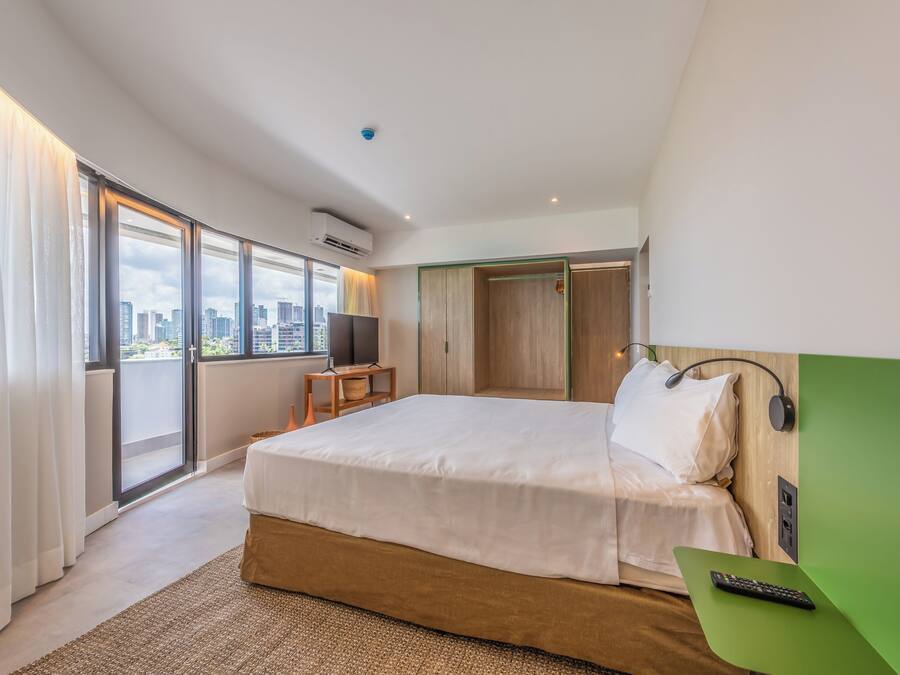 Quarto de hotel moderno com uma cama grande, uma televisão, uma escrivaninha verde e vista para a cidade pela janela.