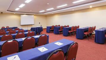 Uma sala de conferência com mesas, cadeiras dispostas em fileiras e uma tela de projeção.