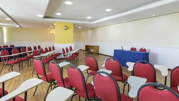 Sala de conferências com fileiras de cadeiras vermelhas e mesas brancas, equipada com luminárias no teto.