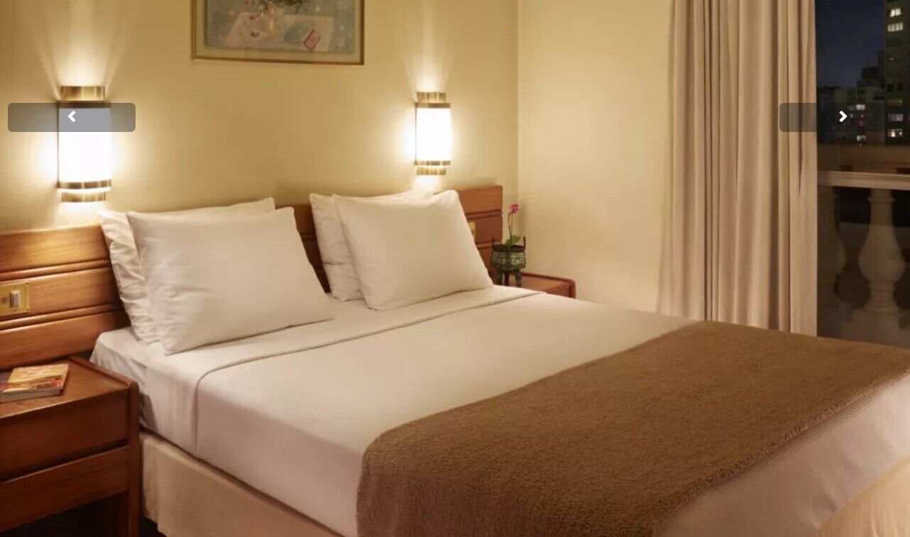 Quarto de hotel com uma cama, duas mesas de cabeceira, um criado-mudo, uma luminária e uma janela com cortinas.