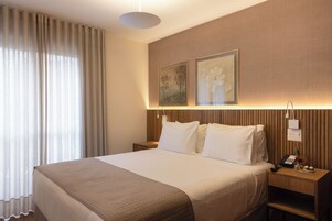 Quarto de hotel com uma cama grande, cabeceira de madeira, duas obras de arte emolduradas, mesa de cabeceira com abajur e criado-mudo com telefone e objetos decorativos.