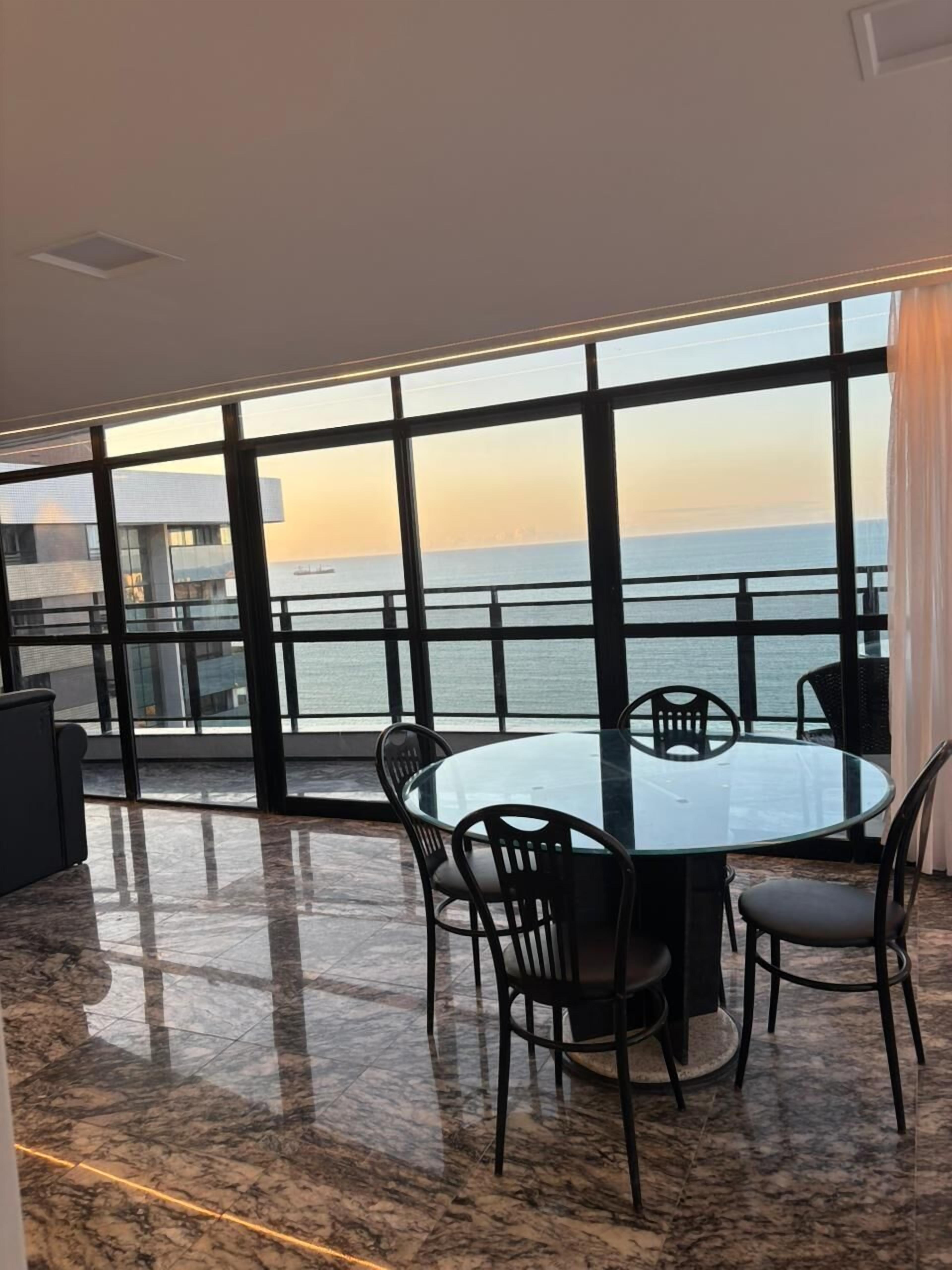 Uma sala de jantar moderna com mesa e cadeiras de vidro, amplas janelas com vista para o mar e piso de mármore.