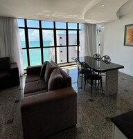 Uma sala de estar moderna com uma grande janela com vista para o oceano, um sofá castanho escuro, uma mesa de jantar com cadeiras e uma moldura na parede.