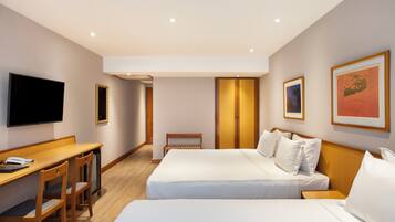 Quarto de hotel com duas camas, uma escrivaninha, uma cadeira, televisão e quadros nas paredes.
