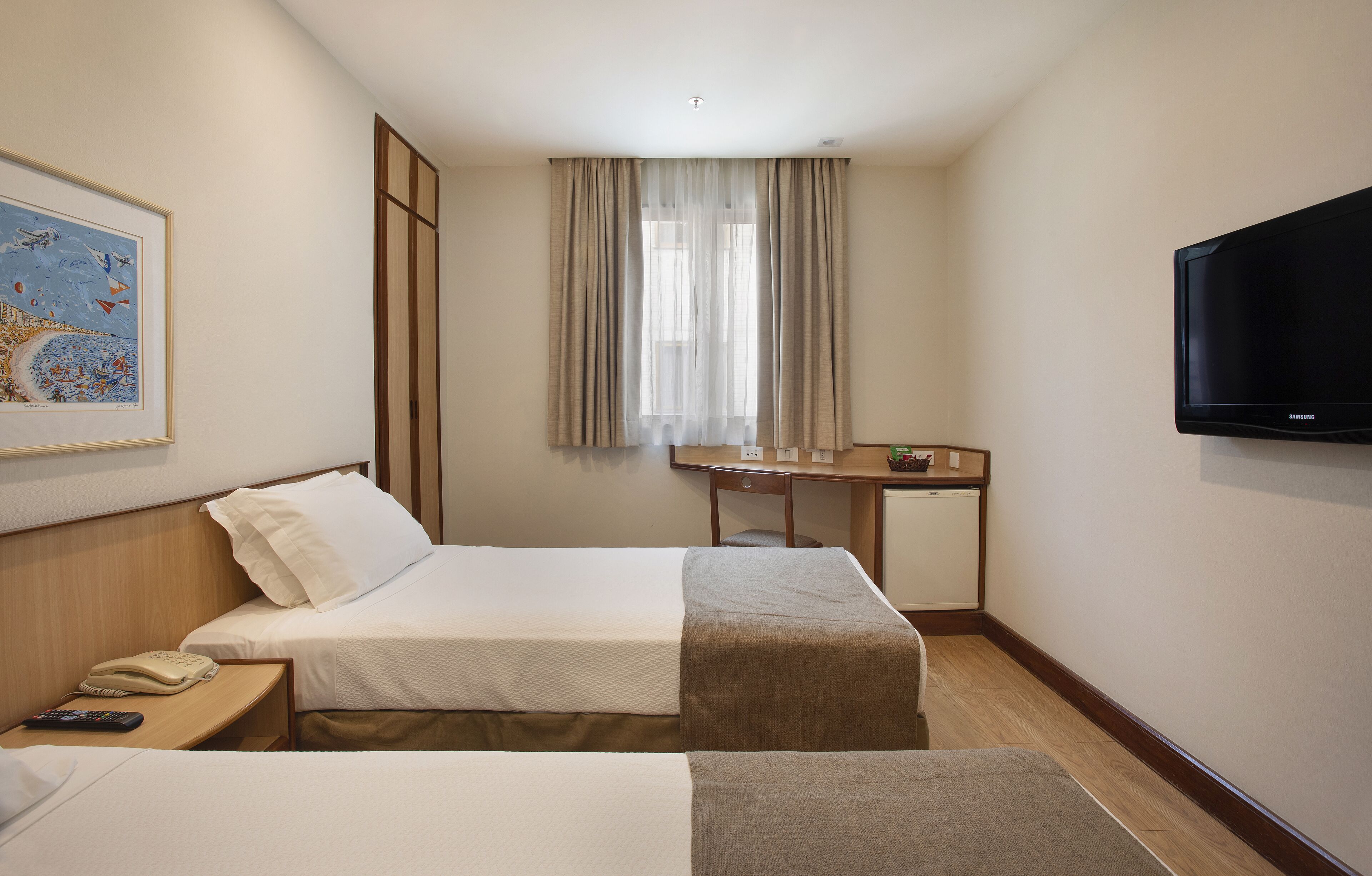 Quarto de hotel com duas camas, uma escrivaninha, uma televisão e um quadro na parede.