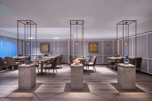 Uma área de refeições moderna com mesas, cadeiras, luminárias pendentes e vasos decorativos em pedestais.