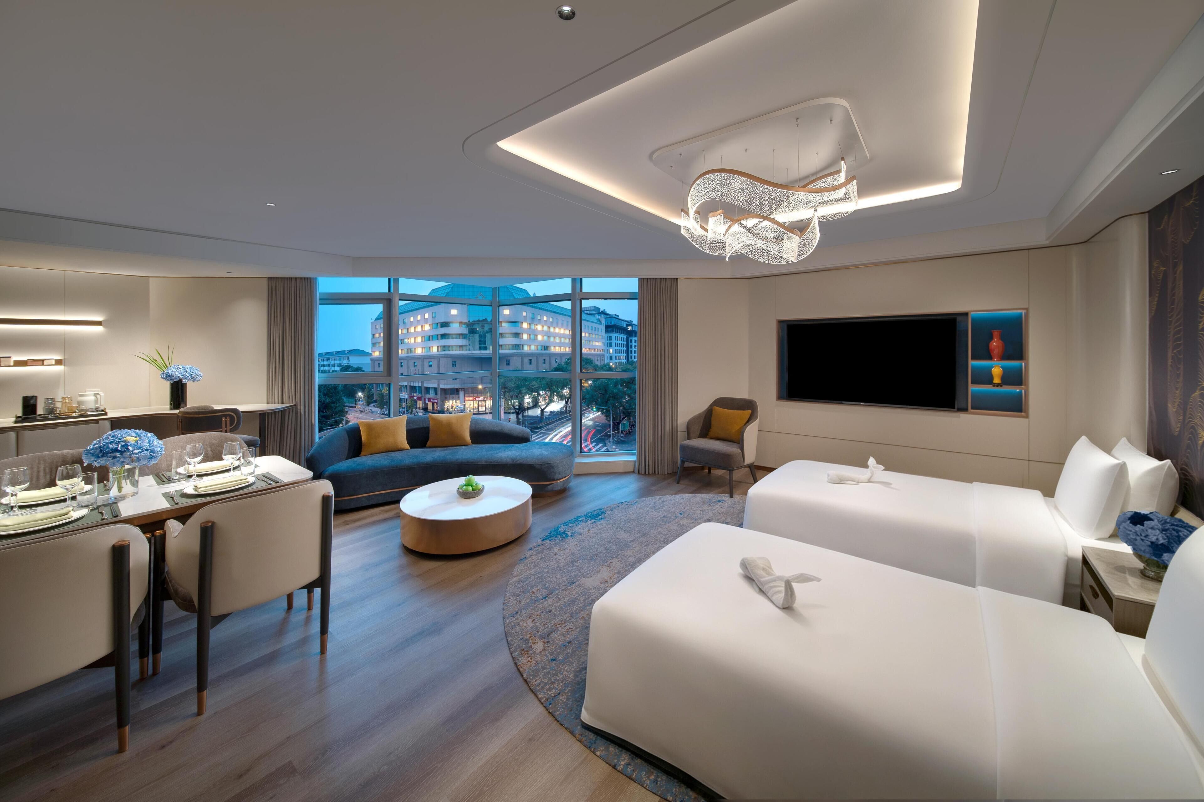 Quarto de hotel moderno com área de refeições, sofá, televisão e uma grande janela com vista para a cidade.