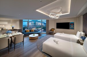 Quarto de hotel moderno com área de refeições, sofá, televisão e uma grande janela com vista para a cidade.