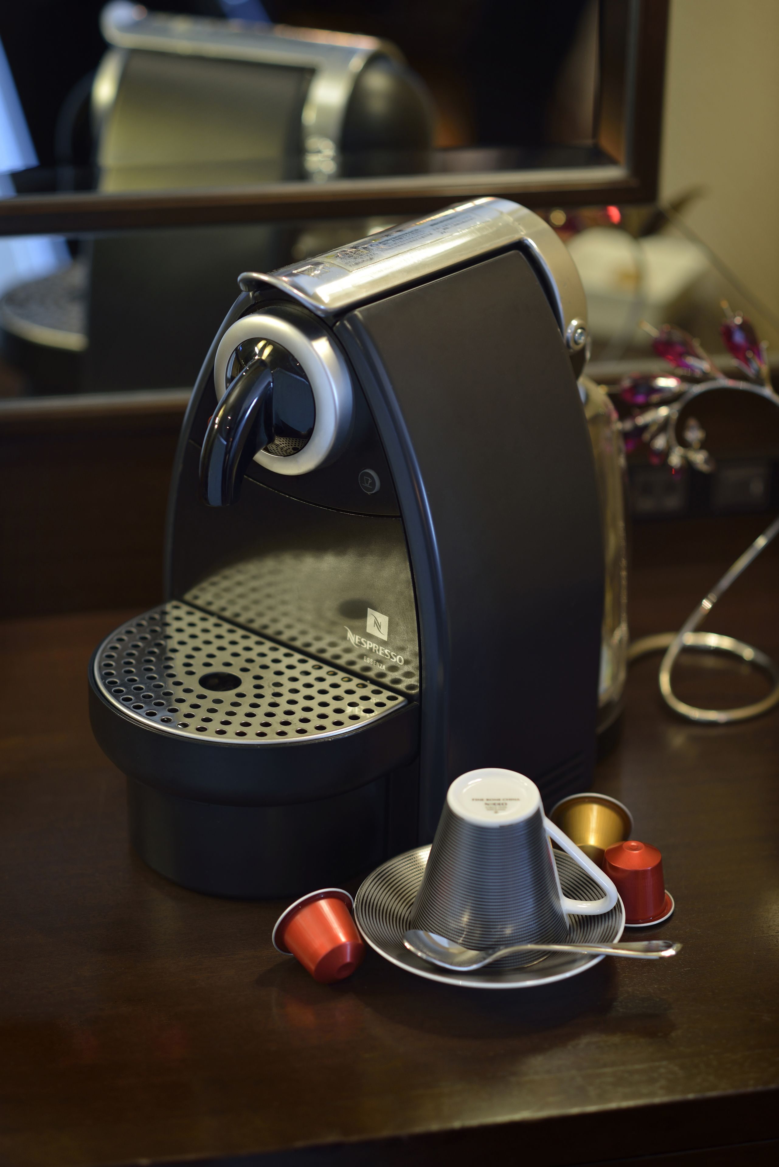 Uma máquina de café Nespresso com uma bandeja coletora, uma xícara de metal e uma colher sobre uma mesa de madeira.