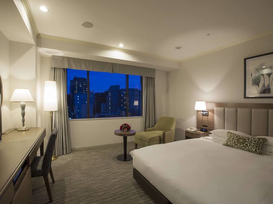 Quarto de hotel com cama, escrivaninha com cadeira, mesinha com abajur, mesa de cabeceira com abajur e janela com vista para a cidade ao entardecer.