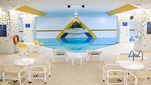 Piscina interna com design geométrico em azulejos, cadeiras de descanso brancas e amarelas, e uma área de estar central.