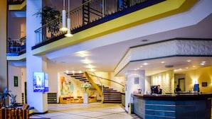 Um hall de hotel com balcão de recepção, escadas e área de estar com plantas.