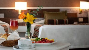 Quarto de hotel com duas camas, uma mesa de café da manhã com uma cesta de pão, um copo de suco, um vaso com flores e um prato de frutas.