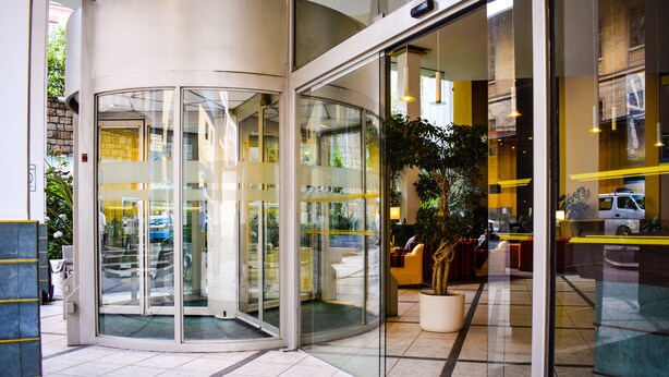 Uma entrada de hotel moderna com porta giratória, paredes de vidro e área de recepção com assentos e uma planta em vaso.