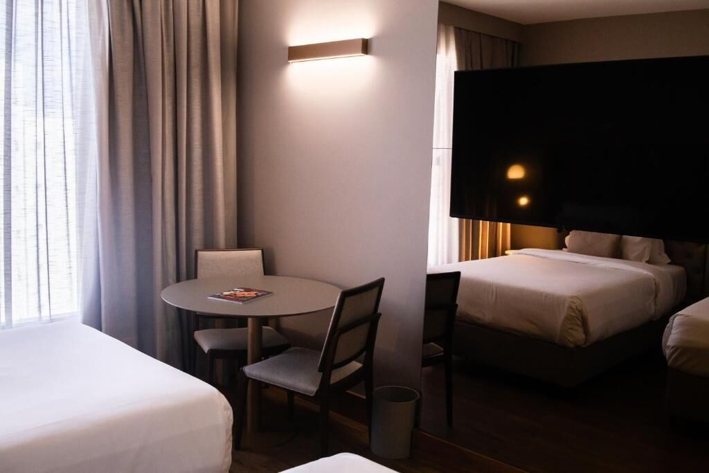 Quarto de hotel com uma cama, televisão, pequena mesa com cadeiras e janela com cortinas.