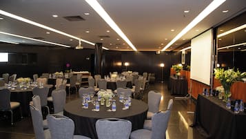 Sala de conferência com mesas redondas, cadeiras cinzas e garrafas de água.