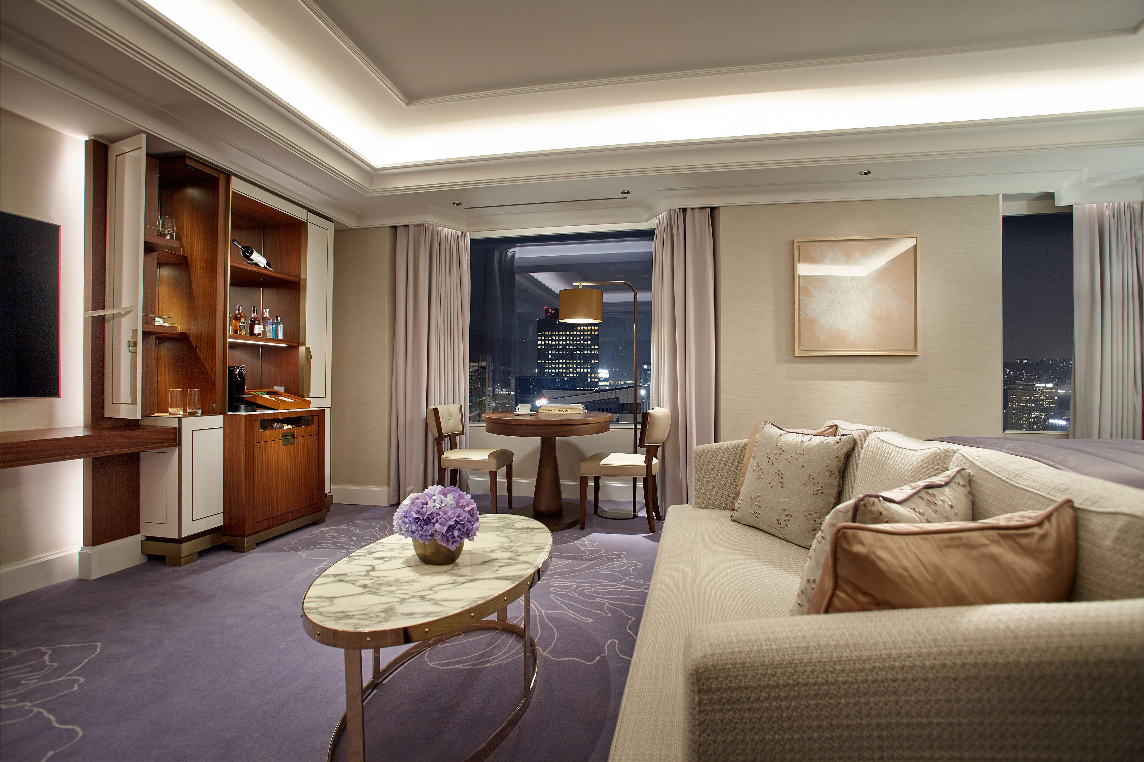 Quarto de hotel com área de estar, minibar, mesa de jantar e vista da cidade à noite.