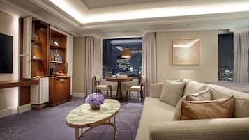Quarto de hotel com área de estar, minibar, mesa de jantar e vista da cidade à noite.