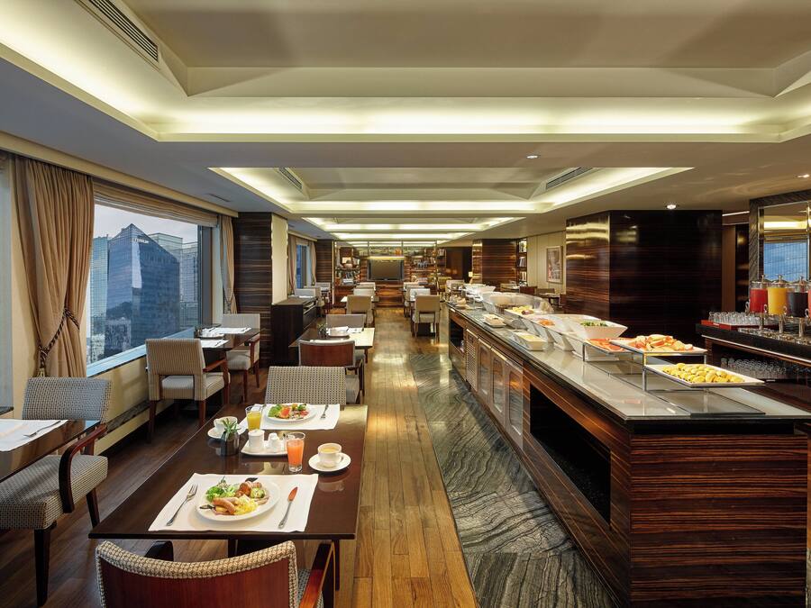 Um espaço de refeições moderno em hotel, com buffet, diferentes opções de assentos e vista para a paisagem urbana.