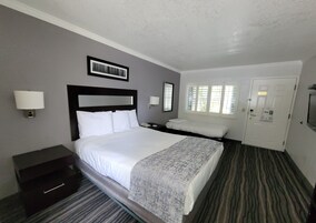 Quarto de hotel com uma cama grande, uma cama menor, uma mesa de cabeceira, uma escrivaninha e uma porta.