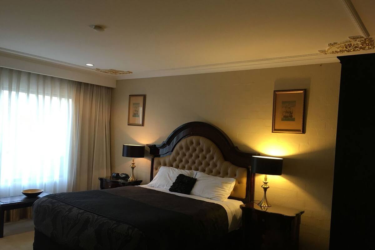 Quarto de hotel com uma cama grande, duas mesas de cabeceira, um criado-mudo, uma janela com cortinas e quadros nas paredes.