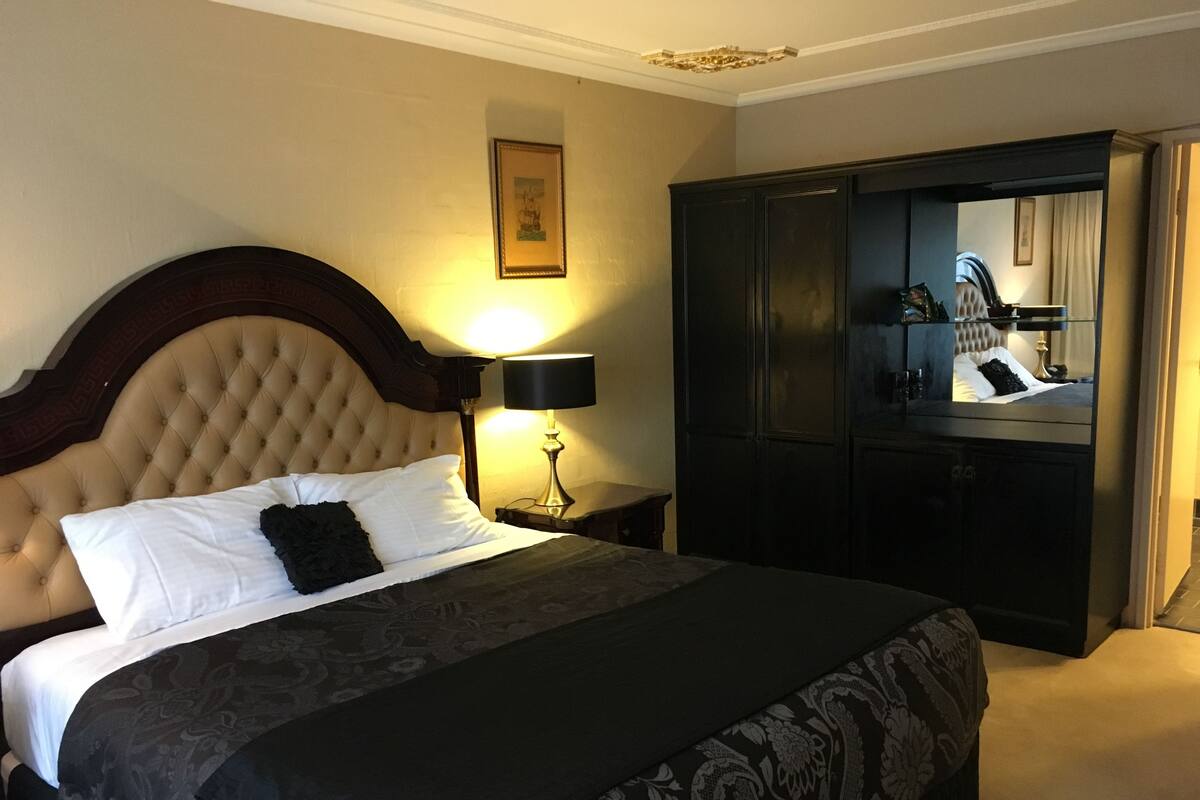 Quarto de hotel com uma cama grande, mesa de cabeceira com abajur, guarda-roupa e espelho.
