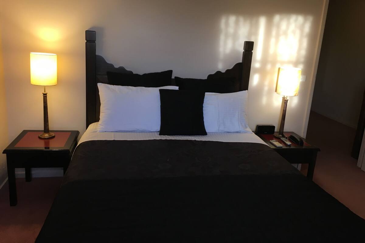 Quarto de hotel com cama, duas luminárias de cabeceira, mesa de cabeceira e cabeceira de madeira.
