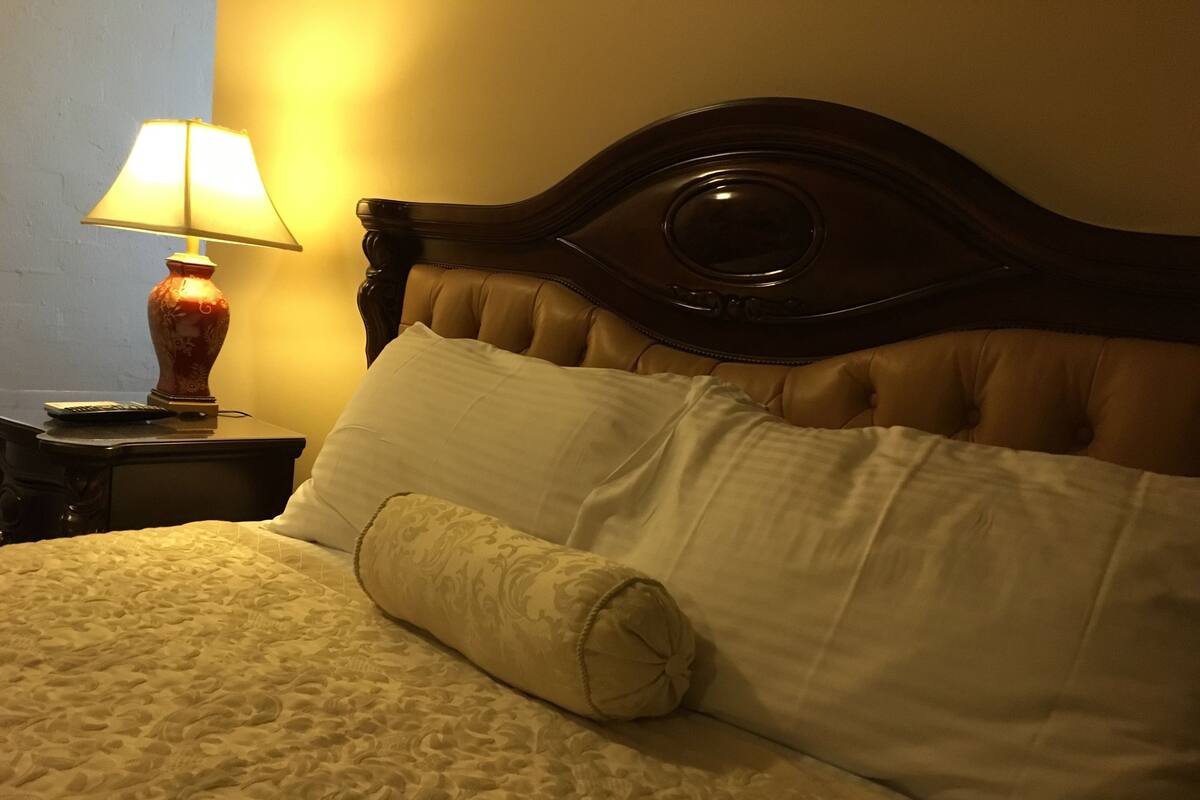Uma cama bem arrumada com cabeceira, abajur e uma mesinha de cabeceira com um controle remoto.