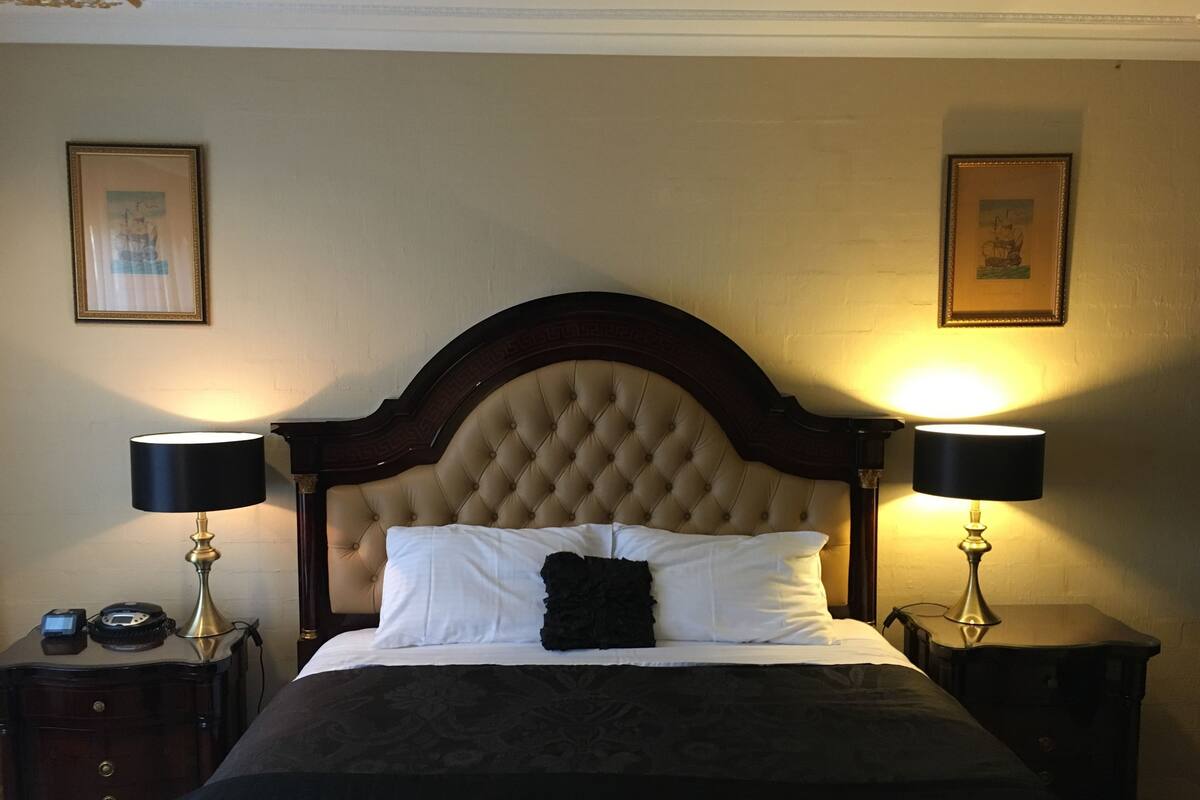 Cama bem arrumada com cabeceira estofada, acompanhada por duas mesas de cabeceira com abajures, em quarto com paredes lisas e quadros enquadrados.