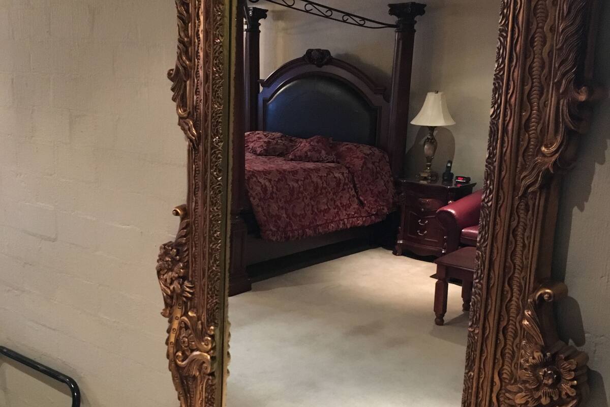 Quarto com um espelho grande e ornamentado, uma cama com cabeceira, uma mesa de cabeceira com abajur e um aparador.
