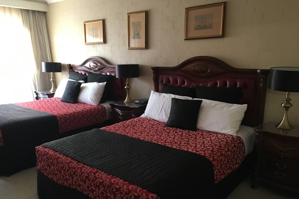 Quarto de hotel com duas camas, cada uma com colcha vermelha e preta, cabeceira de madeira e mesa de cabeceira com abajur.