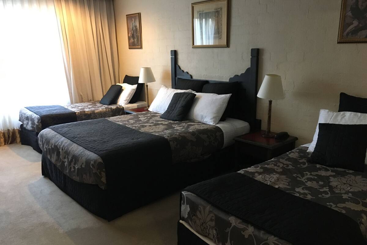 Quarto de hotel com três camas, cada uma com colcha escura estampada e almofadas brancas. Há duas mesas de cabeceira com abajures, e o quarto tem uma paleta de cores neutras com um tapete claro.