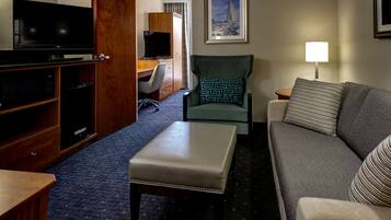 Quarto de hotel com televisão, escrivaninha, poltrona verde, sofá cinzento e luminária.