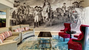 Um saguão com um grande mural histórico, um conjunto de sofás, uma mesa de centro e poltronas estofadas em vermelho.