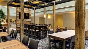 Uma sala de jantar de hotel com mesas e cadeiras de madeira, parede de destaque em pedra e cozinha ao fundo.