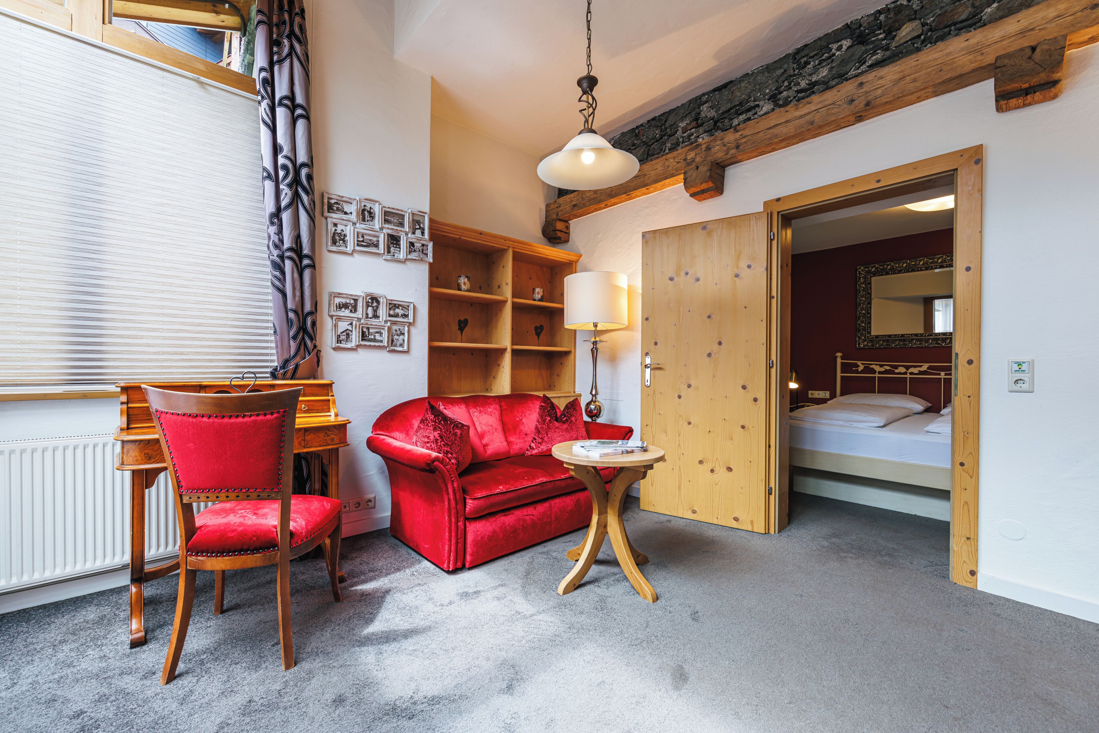 Quarto aconchegante com sofá vermelho, cadeira de madeira e uma pequena mesa. Há uma estante com fotos, uma luminária e uma cama com cabeceira de madeira.
