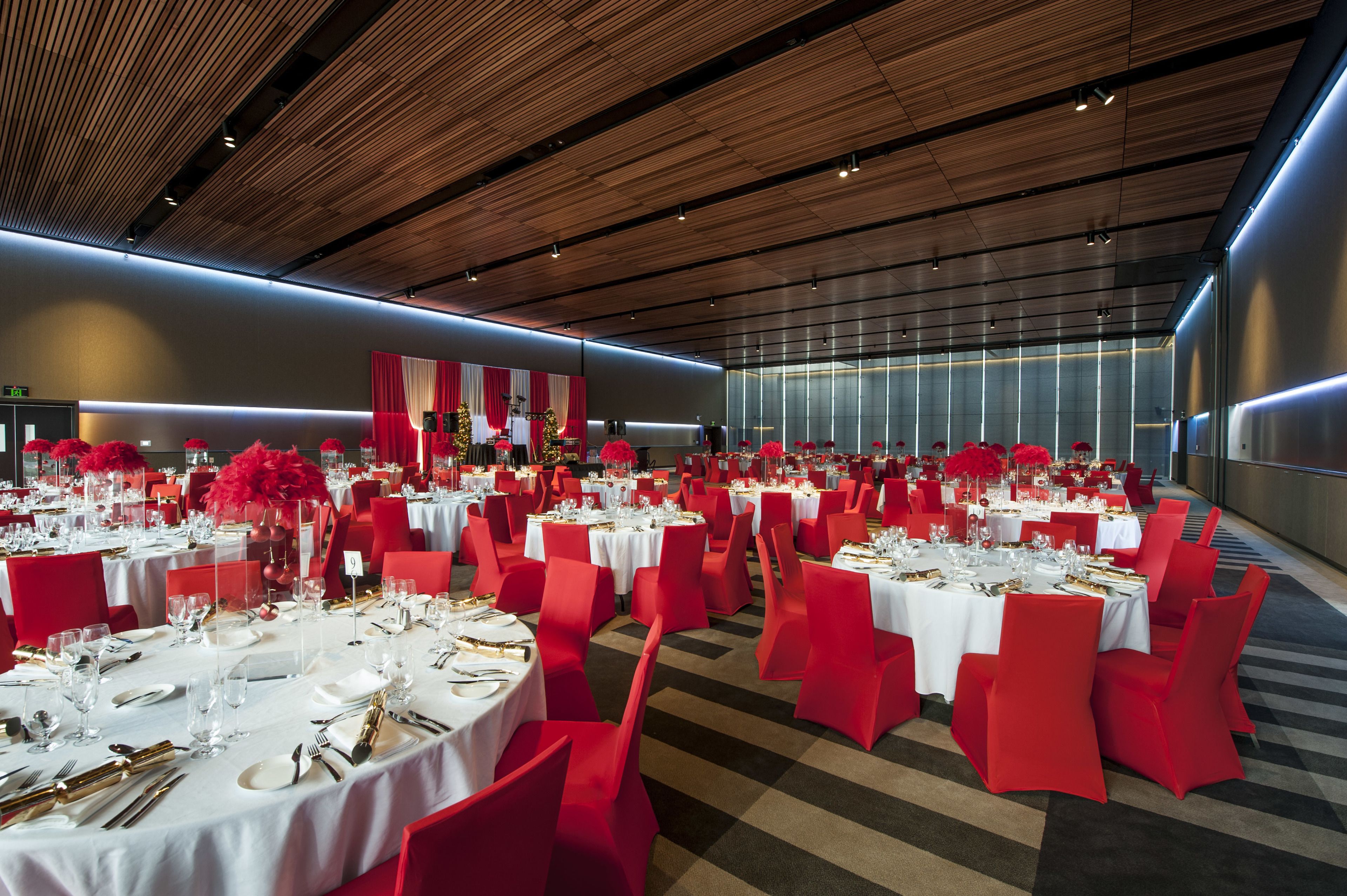 Uma sala de banquetes com mesas redondas preparadas para refeições, cadeiras vermelhas e um design moderno no teto.