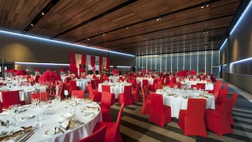 Uma sala de banquetes com mesas redondas preparadas para refeições, cadeiras vermelhas e um design moderno no teto.