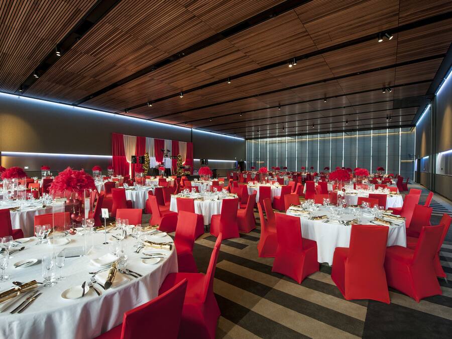 Uma sala de banquetes com mesas redondas preparadas para refeições, cadeiras vermelhas e um design moderno no teto.
