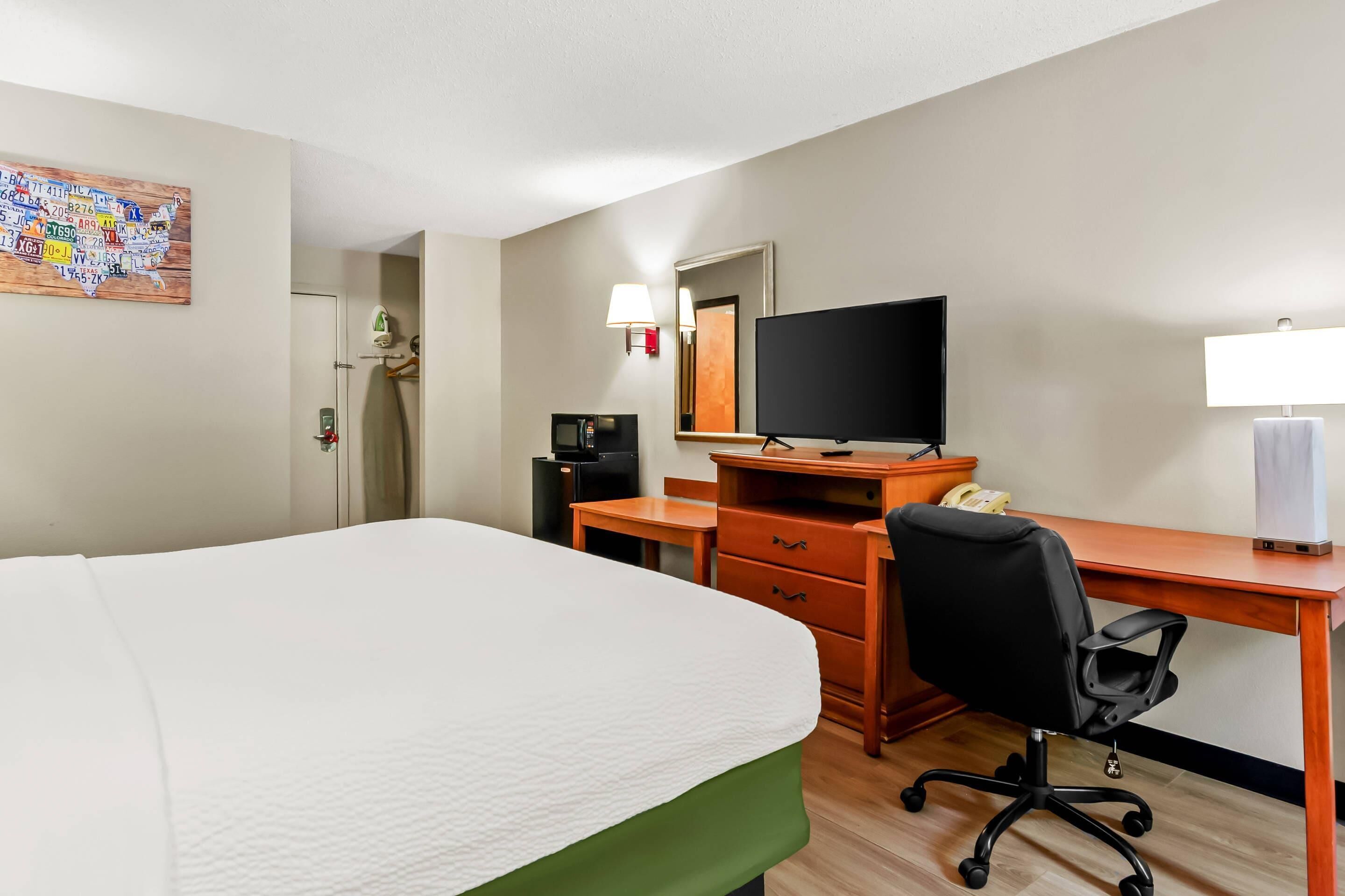 Quarto de hotel com cama, escrivaninha com computador, cadeira, televisão e abajur.