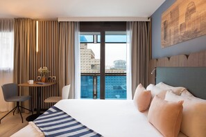 Quarto de hotel com cama, escrivaninha, cadeira, vista da cidade através de uma grande janela e uma pintura de paisagem urbana na parede.